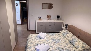 Quarto com cama, mesa de cabeceira, abajur, cômoda e porta para outro cômodo.