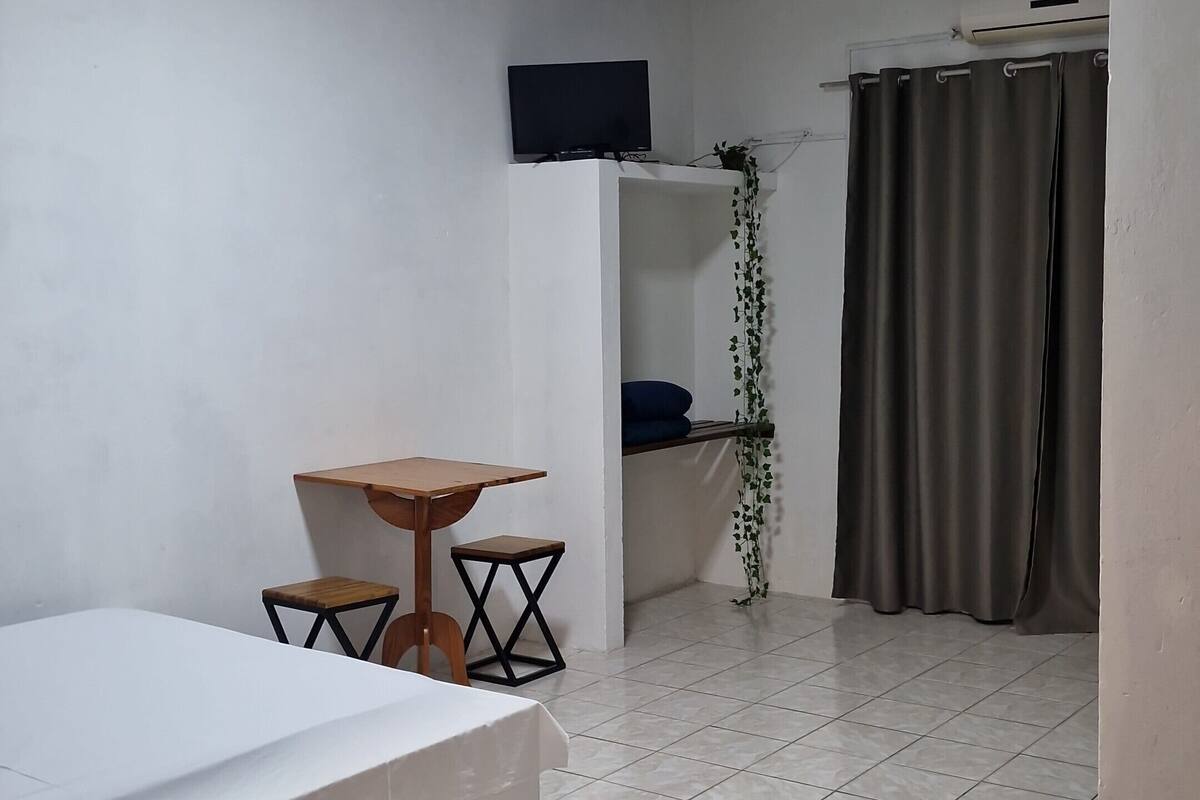 Quarto com chão em azulejo, uma mesa pequena com duas cadeiras, televisão fixada na parede, luminária no teto e cortina.