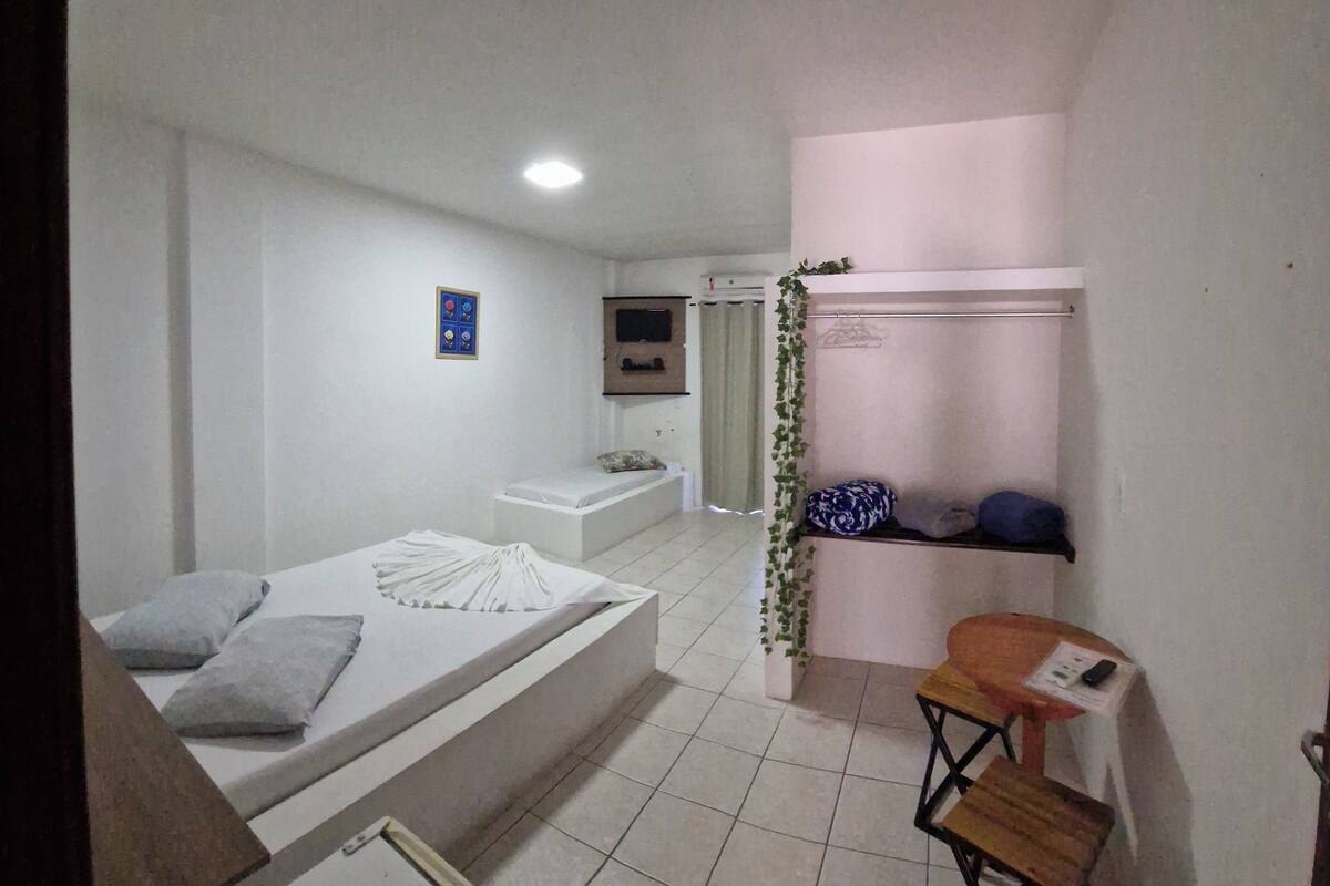 Quarto com cama, uma mesa pequena, cadeira, televisão e chuveiro.