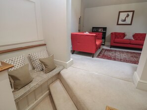 Uma sala de estar com um sofá vermelho, uma poltrona vermelha e um banco estampado. Há uma estante de livros e uma moldura com uma imagem na parede.
