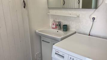Casa de banho compacta com lavatório branco, espelho, máquina de lavar roupa e uma pequena prateleira.