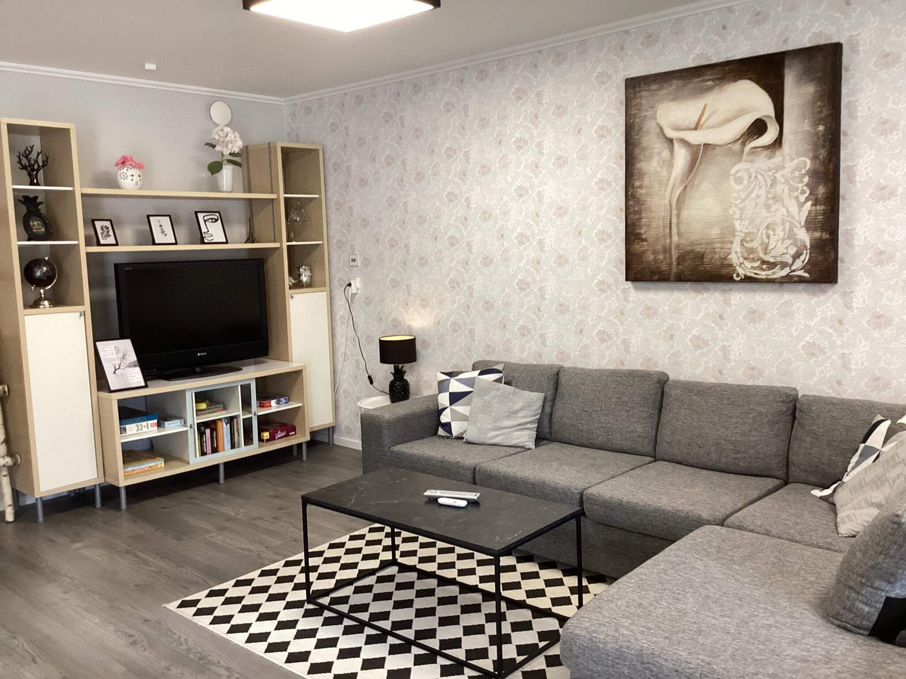 Uma sala de estar com sofá cinza, mesa de centro preta, tapete xadrez, televisão sobre um suporte de madeira e uma parede com padrão floral.