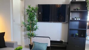 Uma sala de estar moderna com uma mesa de centro preta, um vaso de planta, um sofá cinza, uma poltrona preta, um tapete escuro, uma TV fixada na parede e uma estante preta.