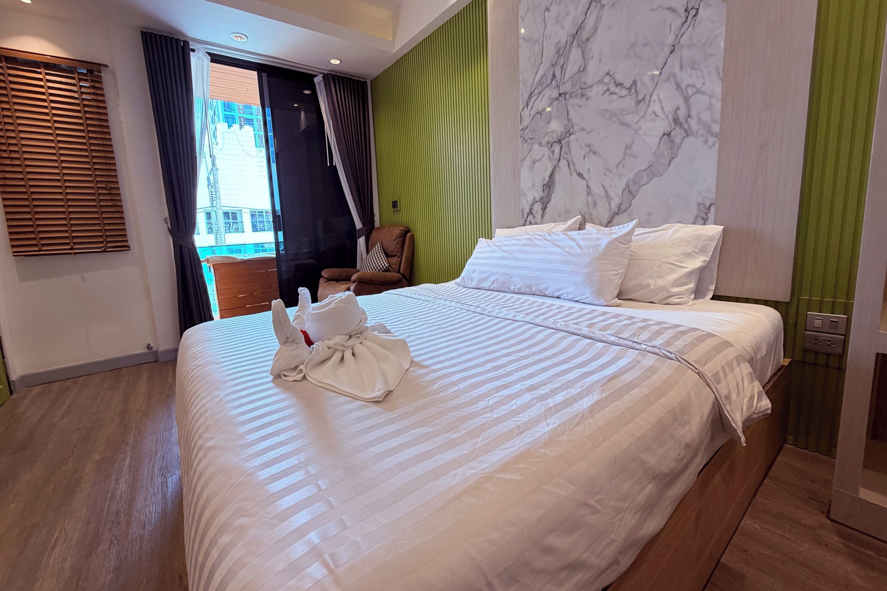 Quarto de hotel com uma cama grande, uma parede de destaque verde e vista para a cidade pela janela.
