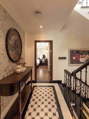 Um corredor com piso estampado que leva a uma sala de jantar, uma escada com corrimão de metal e uma parede com papel de parede floral e um quadro emoldurado.