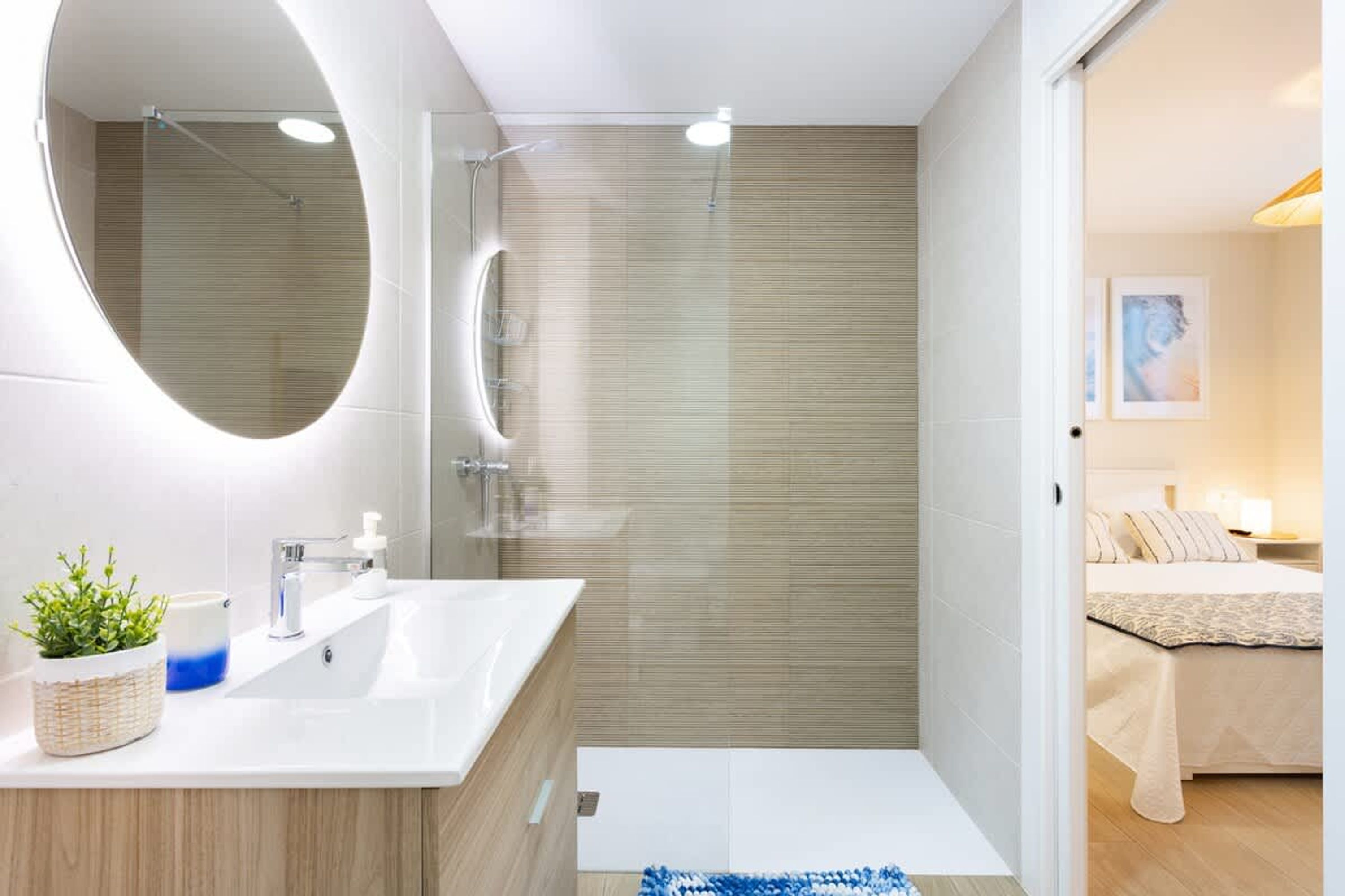 Uma casa de banho moderna com um espelho grande, uma pia, um chuveiro e um quarto visível com uma cama e uma mesa de cabeceira.