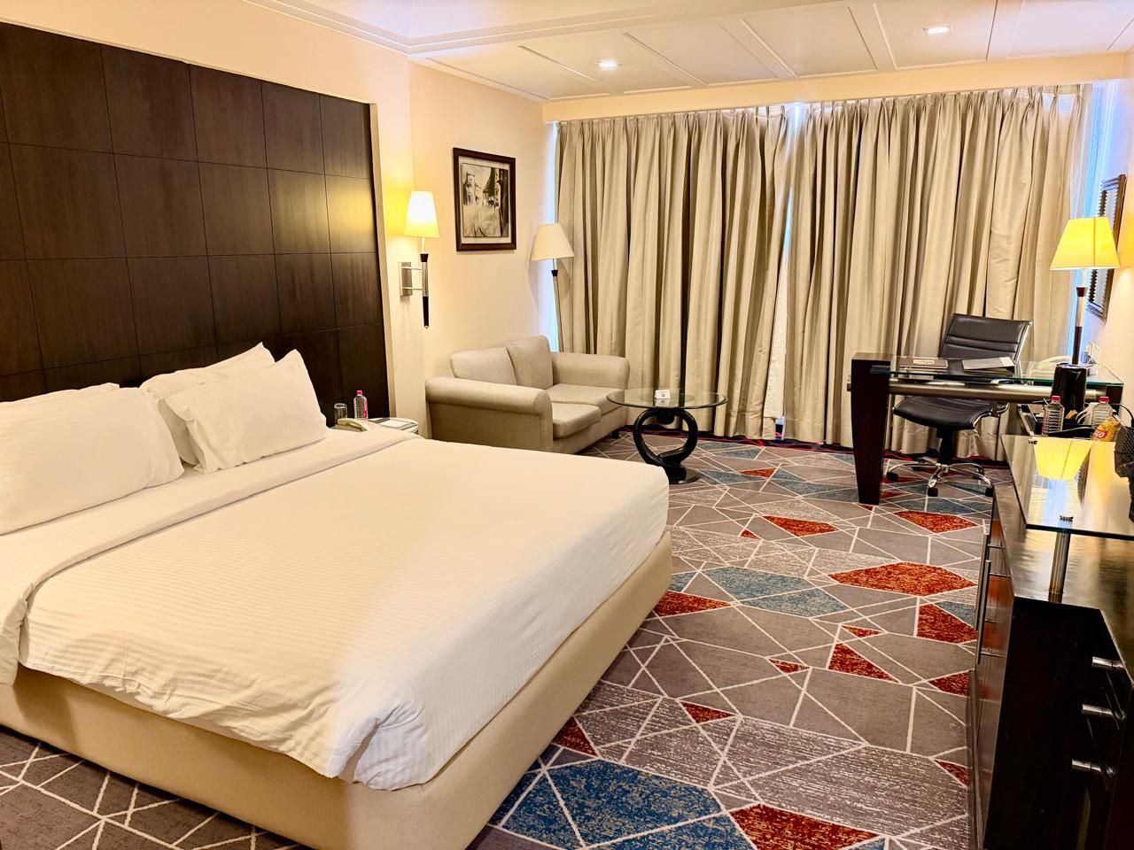 Quarto de hotel com uma cama grande, escrivaninha, cadeira e tapete estampado.