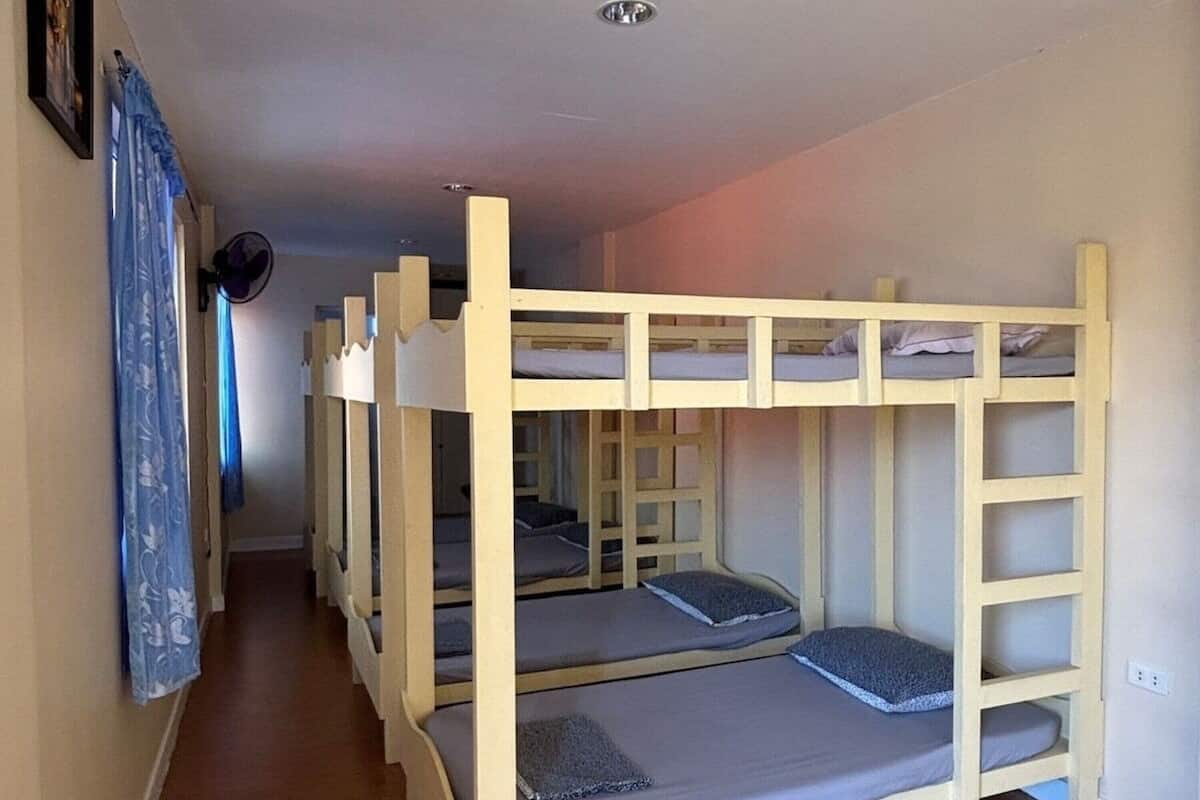 Quarto com beliche, chão de madeira, ventilador de teto e corredor com cortina.