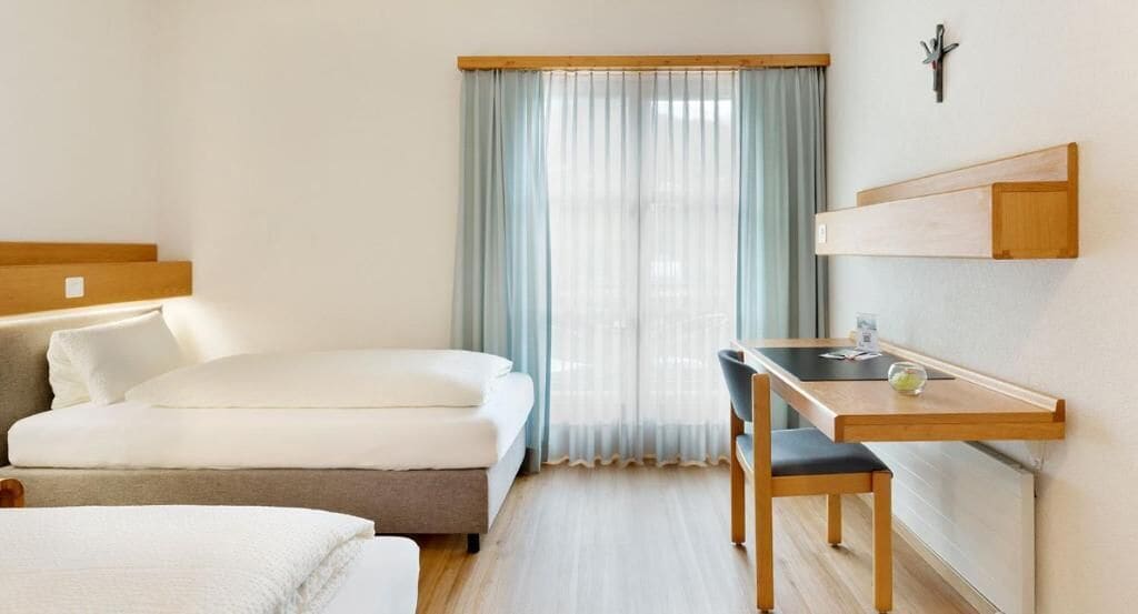 Quarto de hotel com cama, escrivaninha e janela com cortinas.