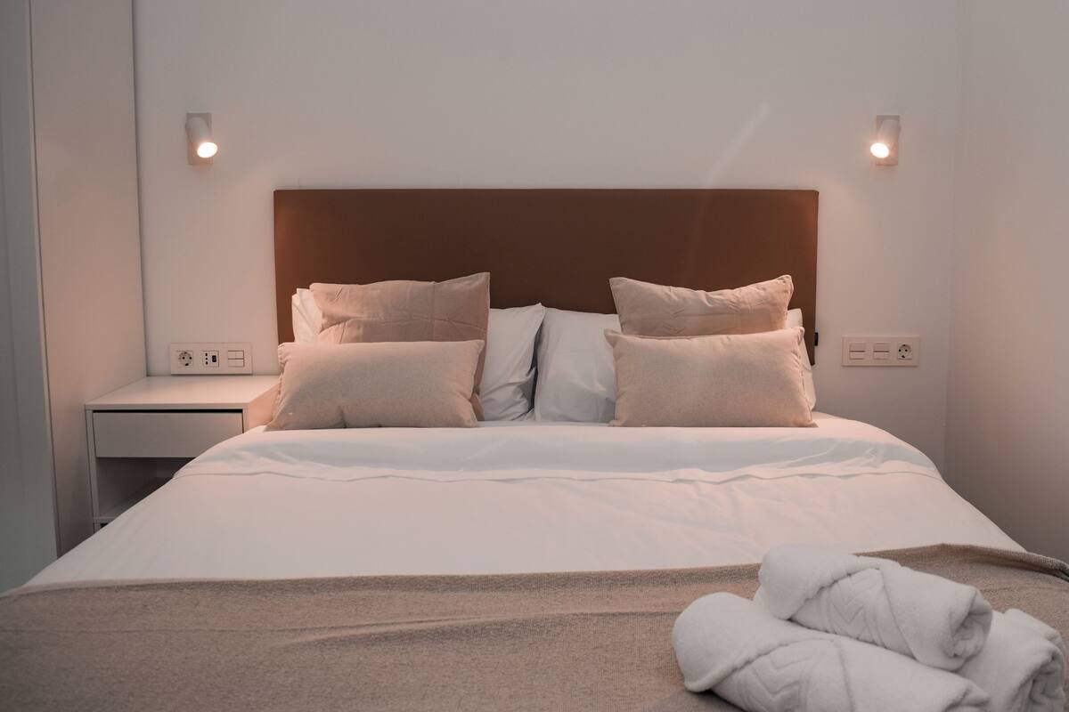 Uma cama bem arrumada com cabeceira, duas mesas de cabeceira e uma tomada na parede.