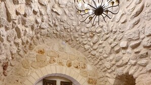Quarto com parede de pedra, porta em arco, cama com roupa de cama branca e um candelabro.