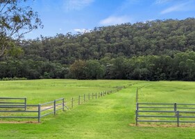 Um campo cercado com céu aberto e uma colina arborizada ao fundo.