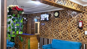 Uma área de recepção com sofá azul, painel de parede de madeira, balcão de recepção, plantas pendentes e um relógio.