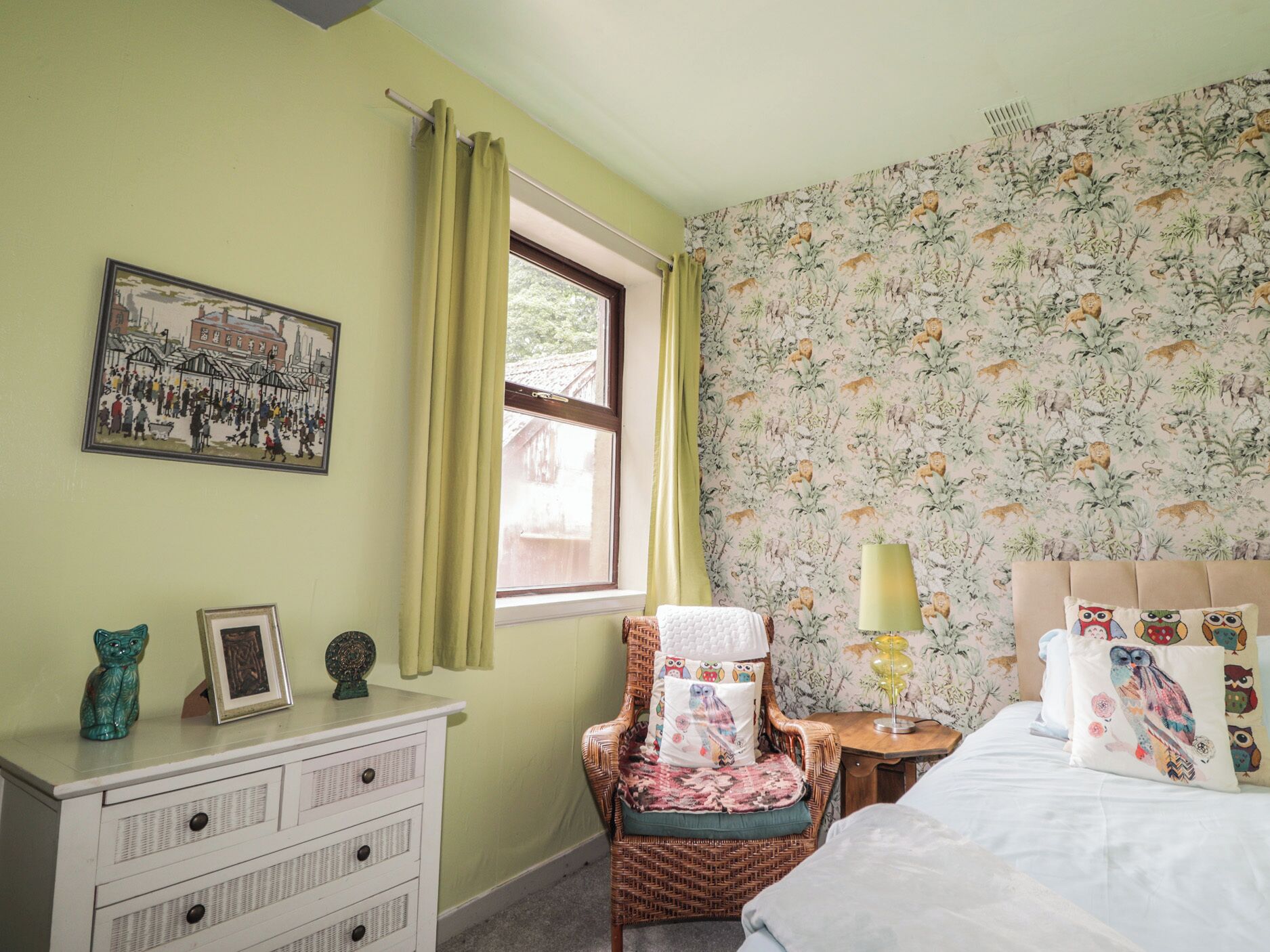 Quarto com cama, cadeira de vime, pequena mesa com abajur, cômoda e janela com cortinas.