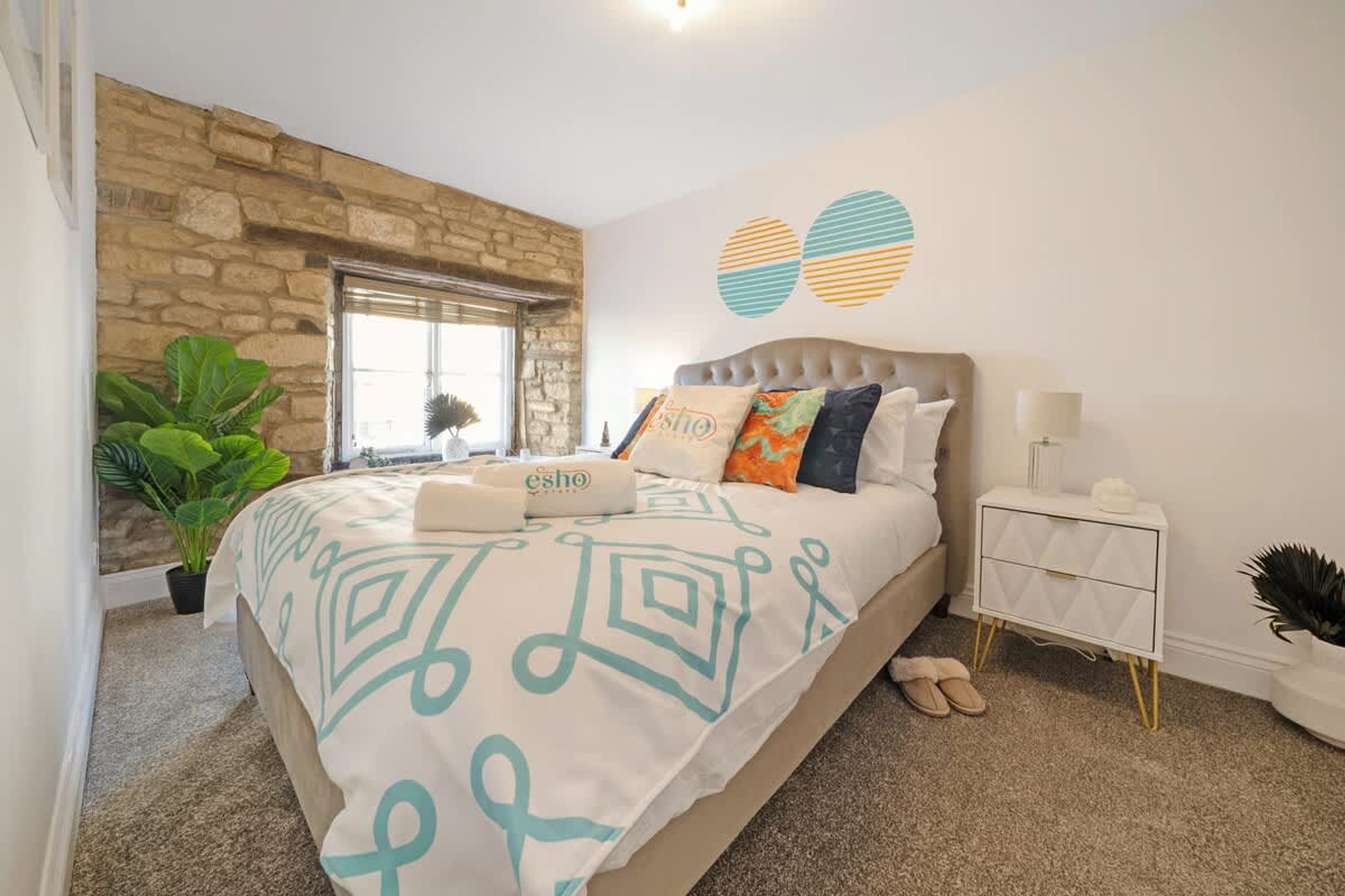 Quarto com parede de pedra, cama com roupa de cama estampada, mesa de cabeceira e planta em vaso.