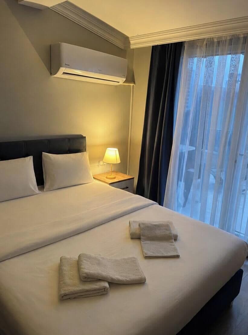 Quarto de hotel com cama, luminária, ar condicionado e janela com cortinas.