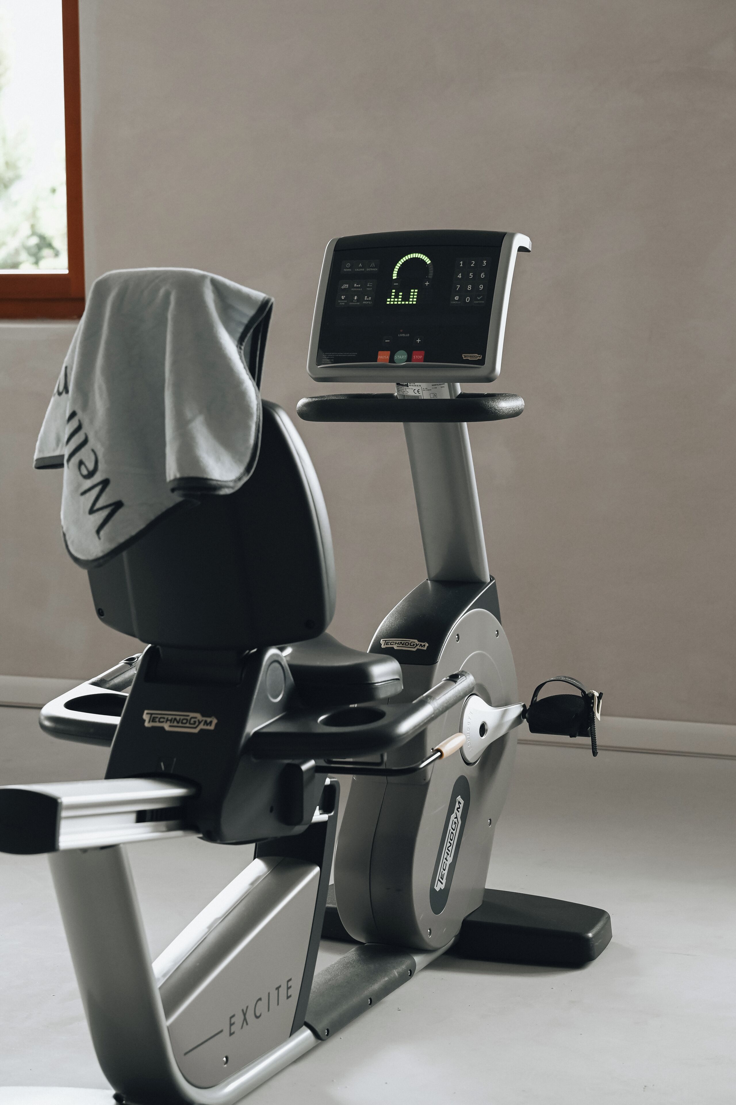 Uma bicicleta ergométrica com mostrador digital, suporte para garrafa de água e a marca 'TECHNOGYM' na estrutura.