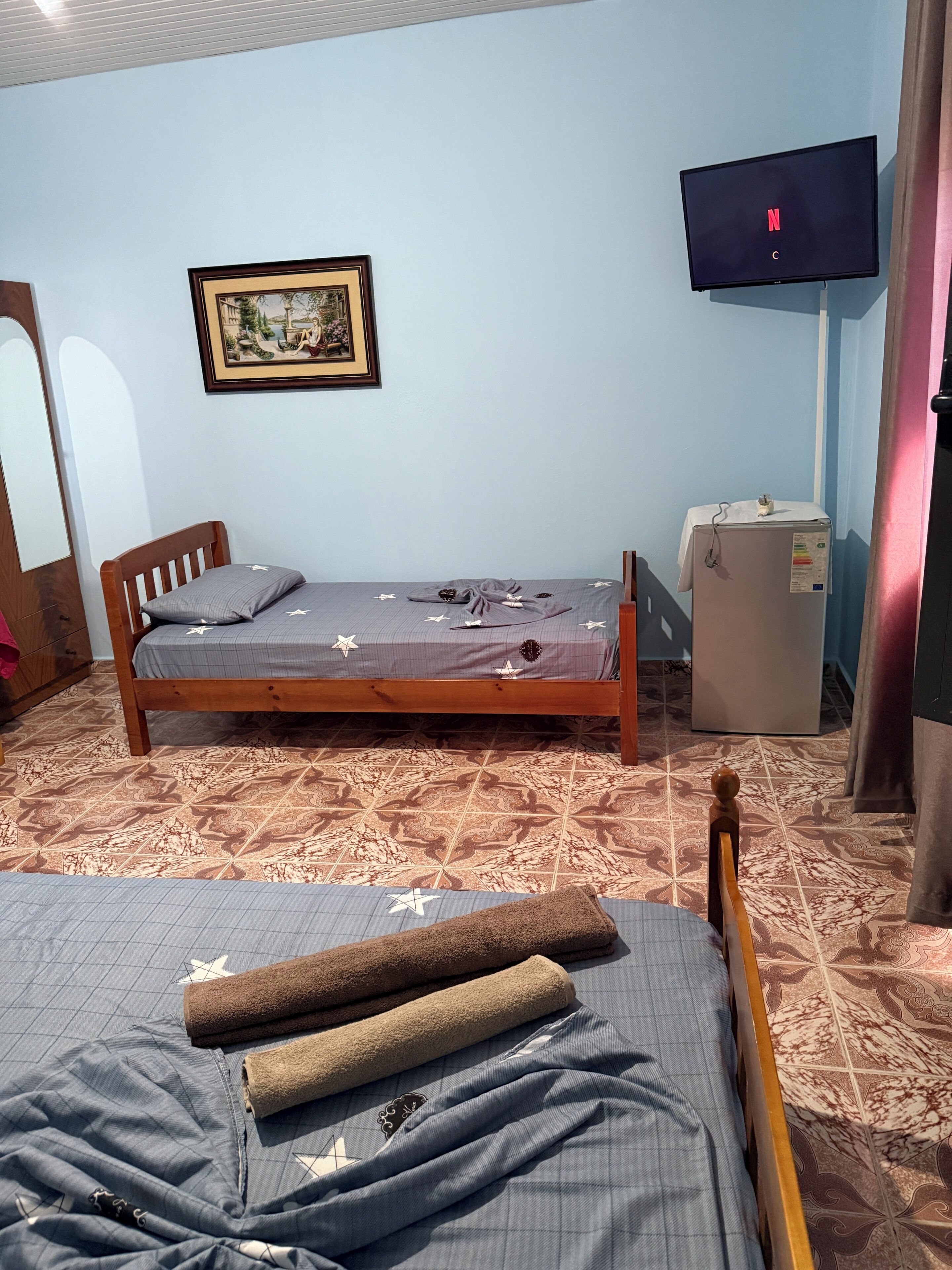 Uma cama de solteiro com estrutura de madeira, uma pequena mesa de cabeceira, uma televisão na parede e um chão com padrão.