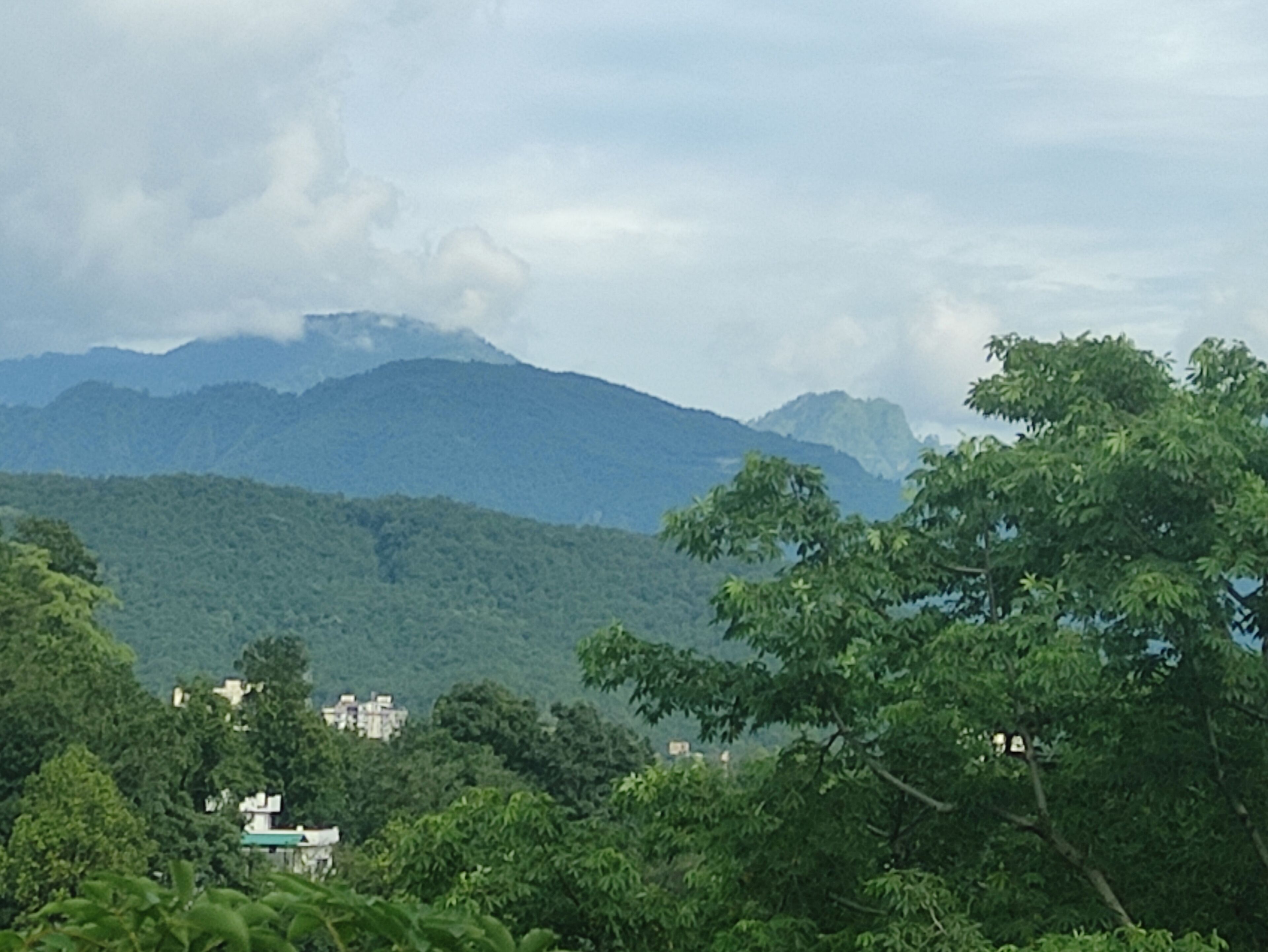 A mountainous landscape with dense greenery and a few buildings in the foreground.