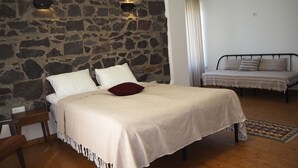 Quarto com parede de pedra, cama com roupa de cama branca, mesa de cabeceira, cadeira de madeira e uma pequena mesa.