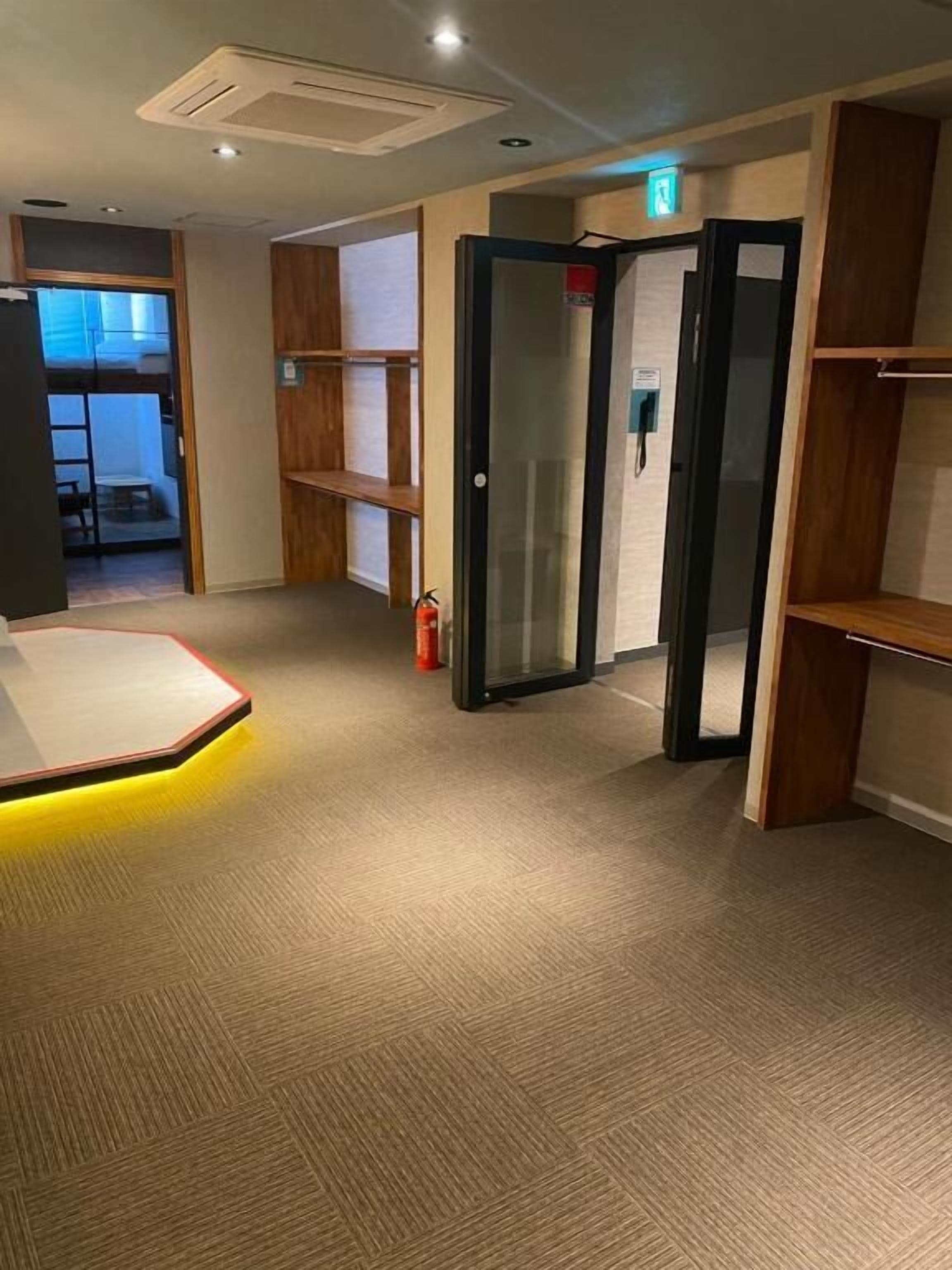 Corredor de hotel com piso carpete, estantes de madeira e porta de vidro com extintor de incêndio.