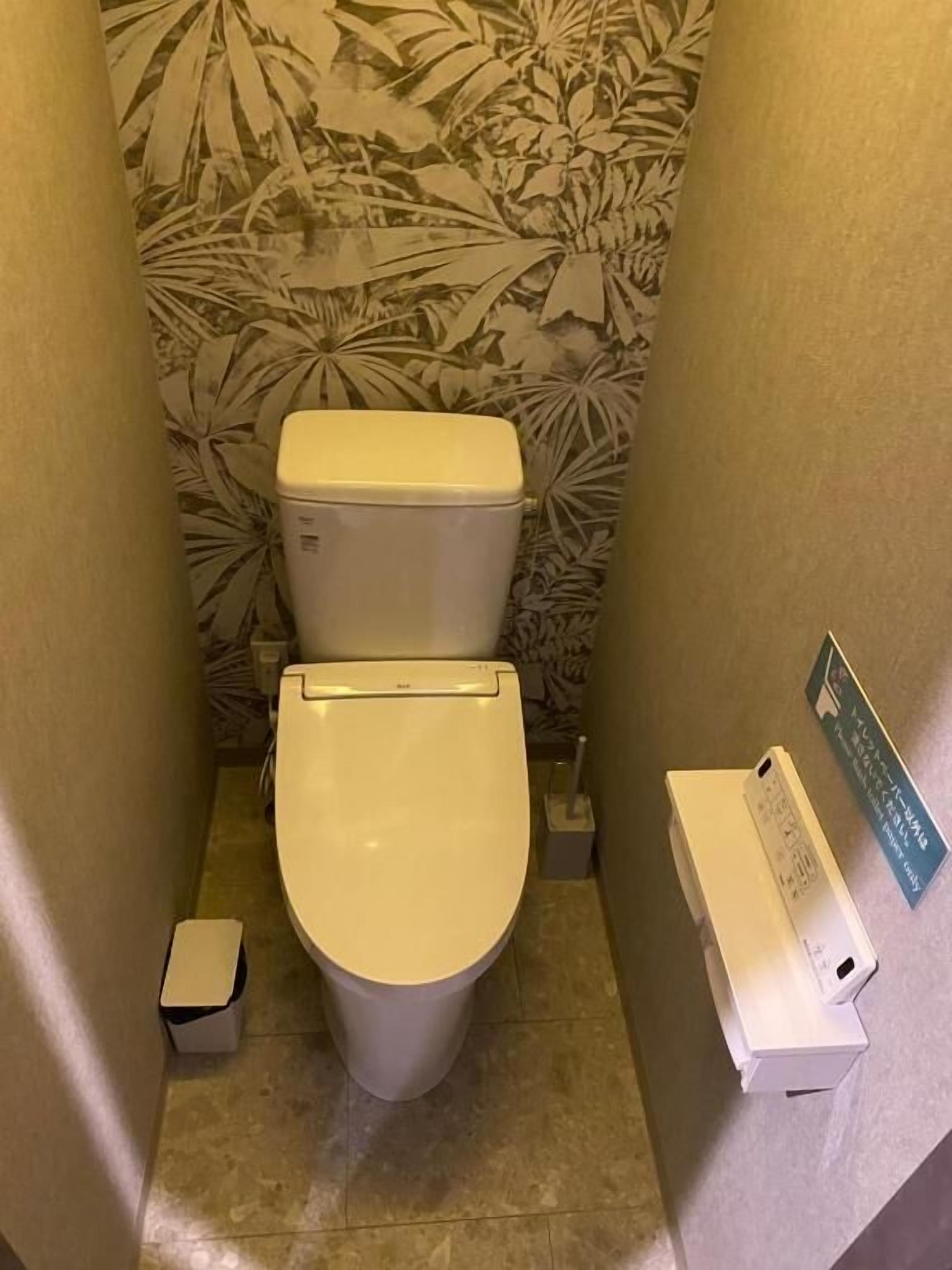 Sanitário com papel de parede floral, uma pequena lixeira e um dispositivo fixado na parede.