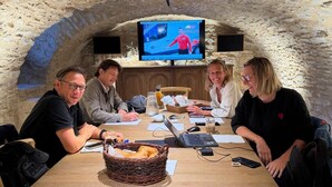 Um quarto com paredes de pedra, uma mesa redonda de madeira, uma televisão e pessoas sentadas ao redor.