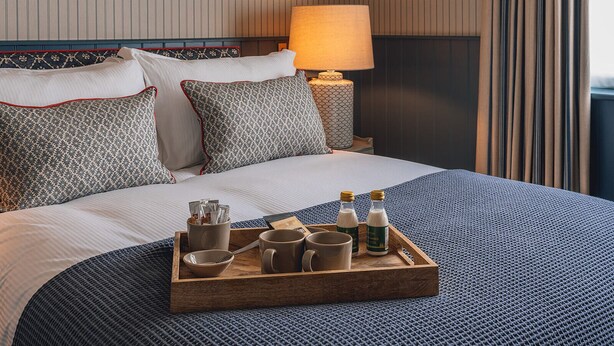 Cama bem arrumada com almofadas estampadas, mesa de cabeceira com abajur e bandeja com itens de café da manhã.