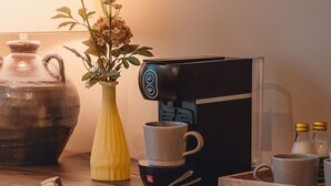 Uma bancada de cozinha com uma cafeteira, um vaso com flores, um bule e uma cadeira.