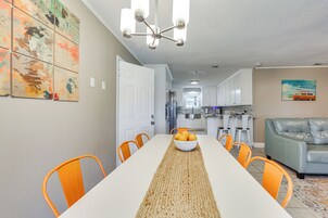 Uma área de refeições com uma mesa comprida, cadeiras laranja, um jogo americano tecido e uma tigela de laranjas.