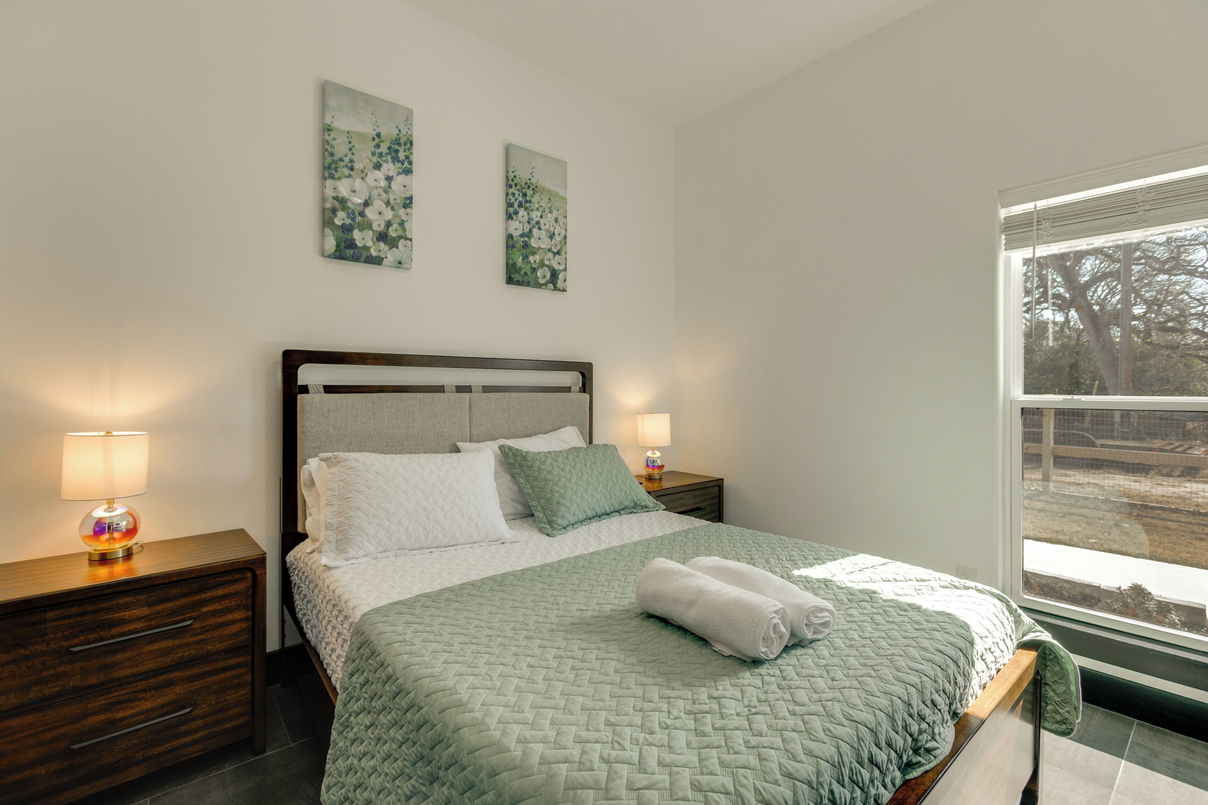Cama bem arrumada com colcha verde, duas almofadas e uma toalha enrolada ao lado. Há duas mesas de cabeceira com luminárias, e o quarto tem uma janela grande com vista para árvores.