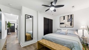 Quarto com cama, ventilador de teto, abajur, banco, espelho e porta.