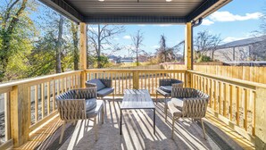 Um terraço ao ar livre com mesa e cadeiras, guarda-corpo de madeira e vista para árvores e casas.