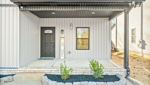 Uma casa com varanda, porta preta, caixa de correio e pequeno jardim com dois arbustos.