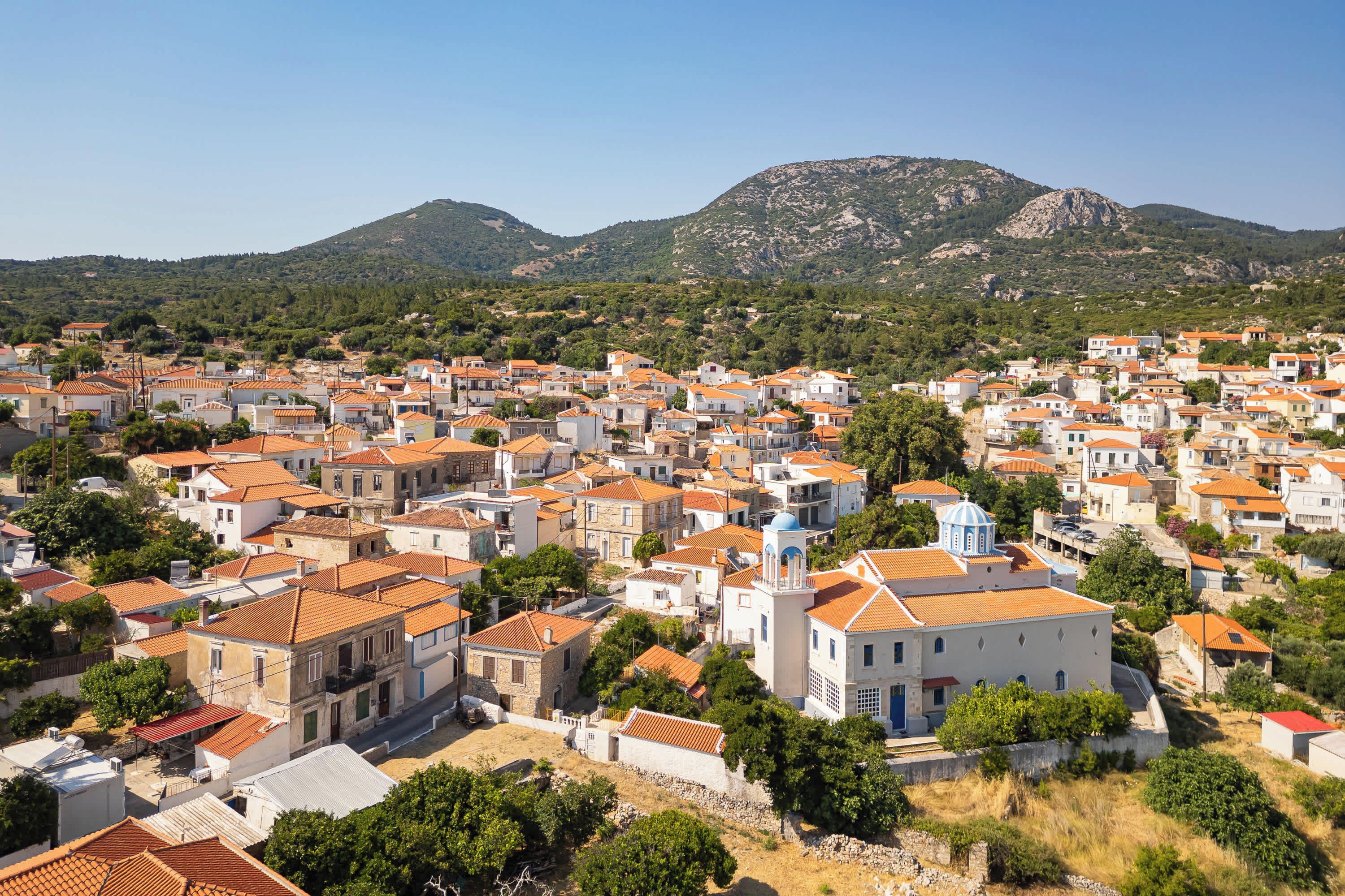 Uma vila costeira com edifícios brancos e telhados vermelhos, rodeada por vegetação e montanhas.