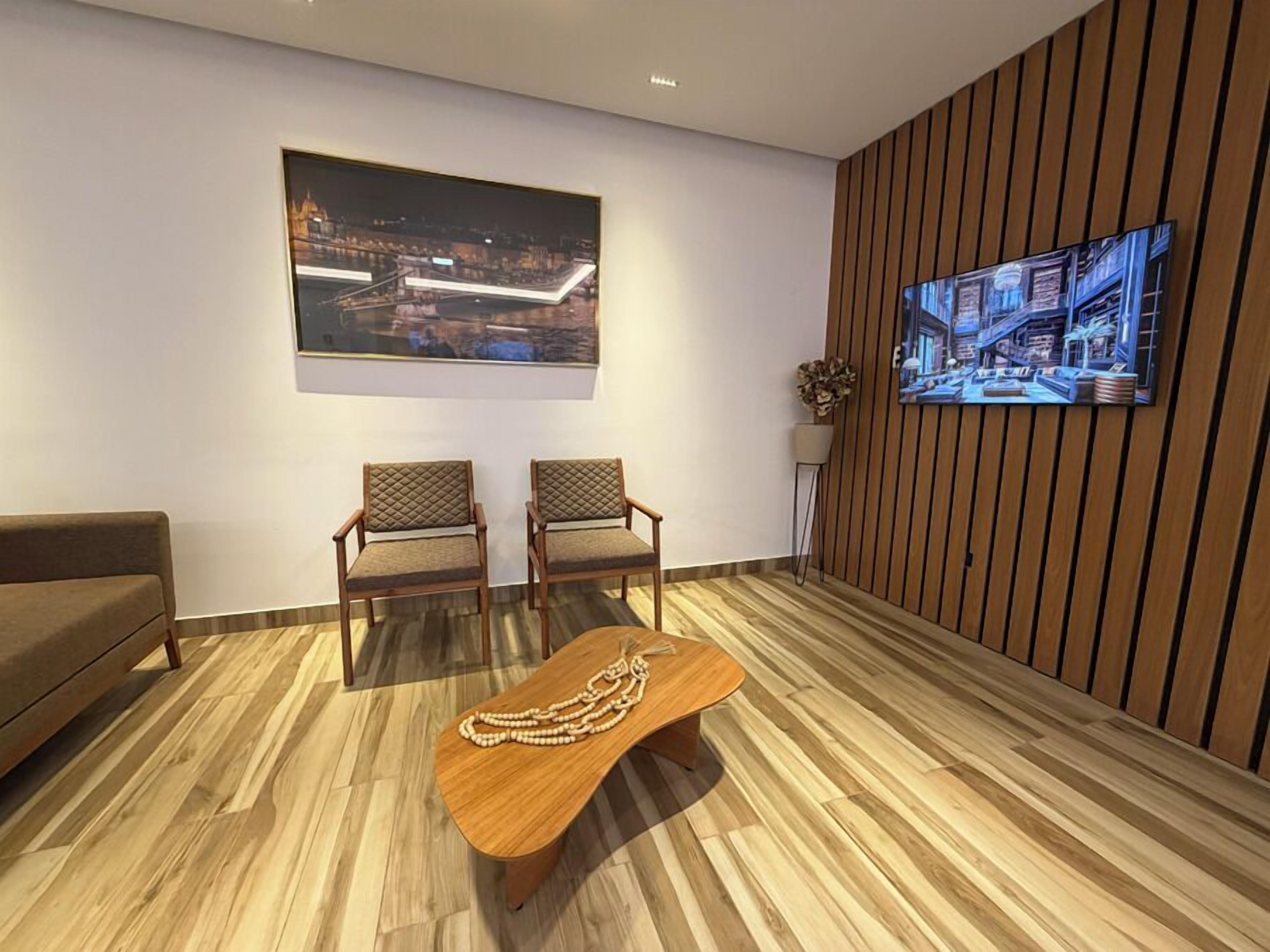 Quarto moderno com piso de madeira, dois poltronas, um sofá, uma mesa de centro de madeira com um colar de contas e uma TV fixada na parede, além de uma grande moldura com uma imagem.