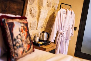Quarto de hotel com parede de pedra, mesa de cabeceira de madeira com chaleira, roupão e almofada decorativa.