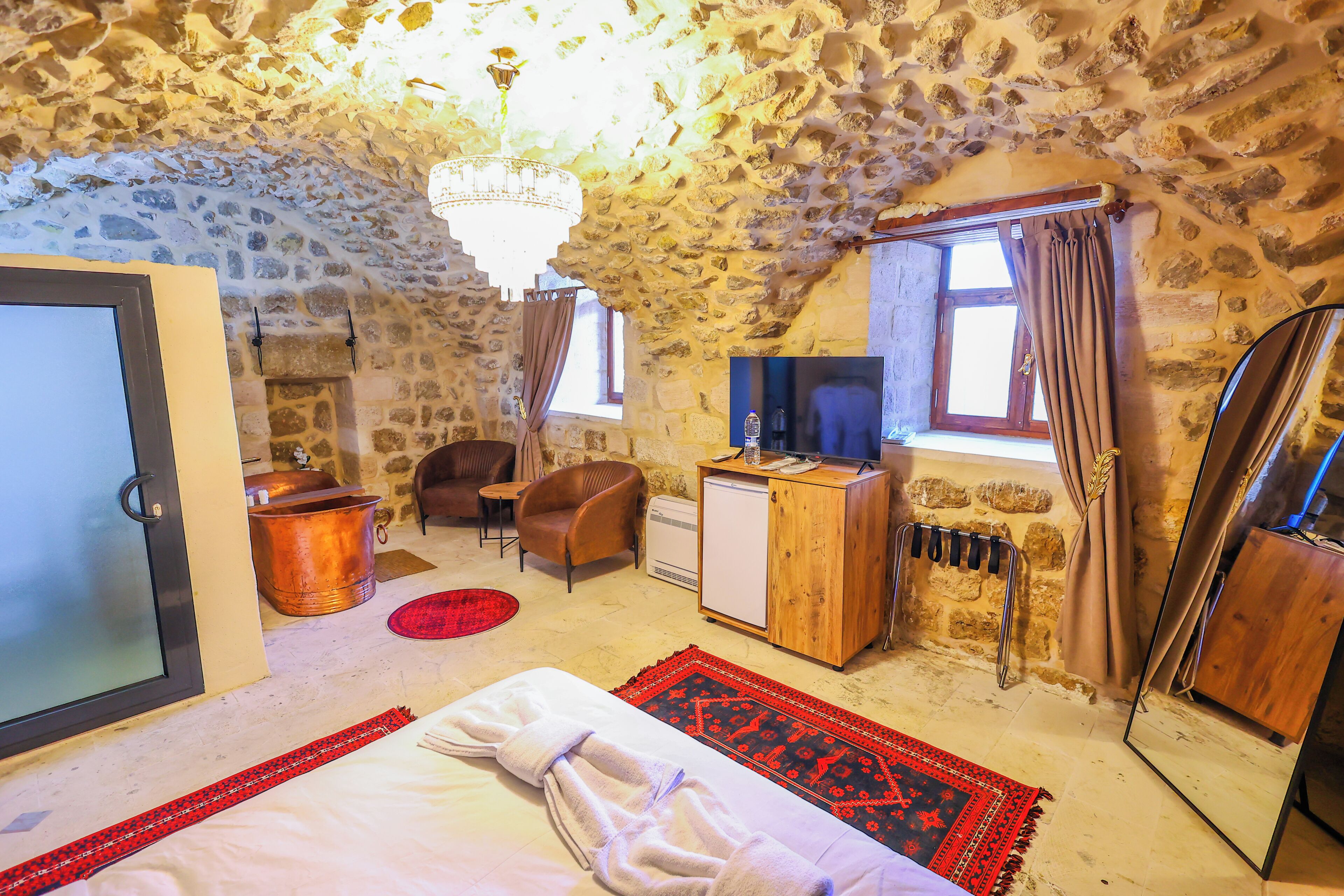 Quarto rústico com teto de pedra, cama com tapete de padrão vermelho e preto, armário de madeira, televisão e uma pequena mesa.