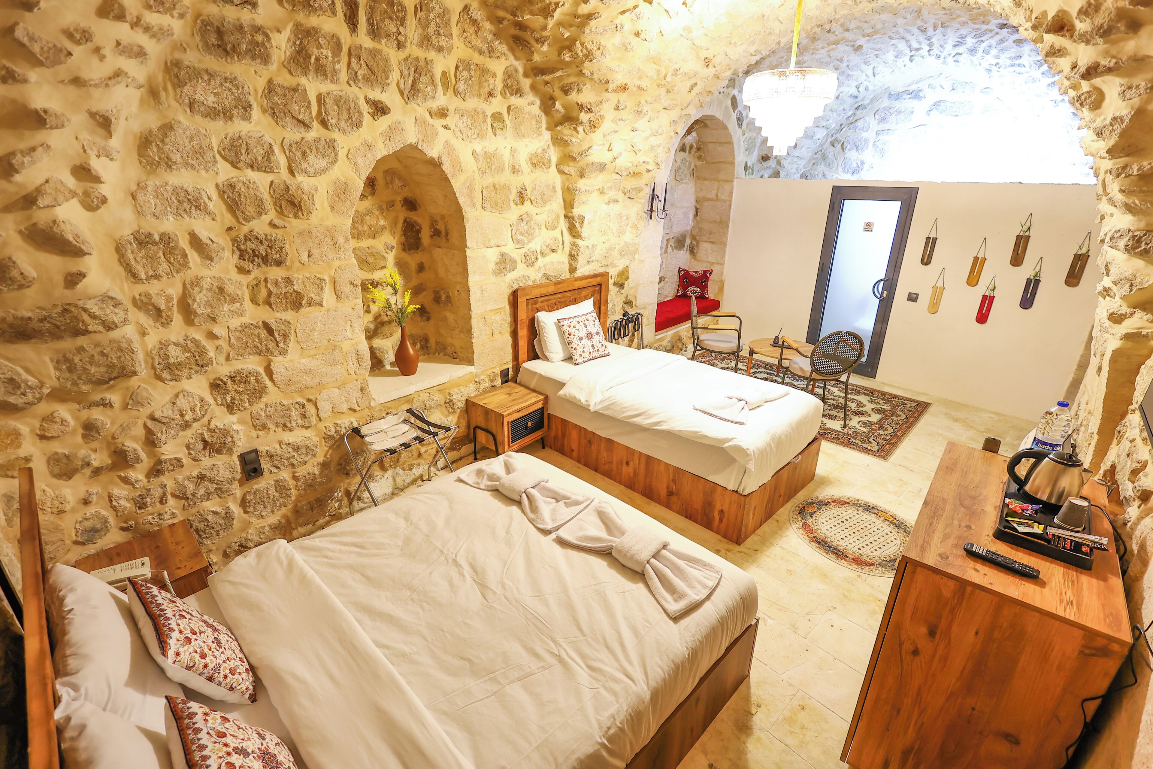 Quarto com paredes de pedra, duas camas, uma mesa de cabeceira de madeira, uma escrivaninha e uma cadeira.