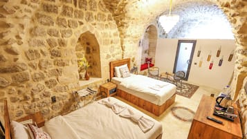 Quarto com paredes de pedra, duas camas, uma mesa de cabeceira de madeira, uma escrivaninha e uma cadeira.