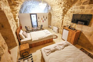 Quarto com paredes de pedra, uma cama, escrivaninha, cadeira, televisão e uma pequena cozinha compacta.