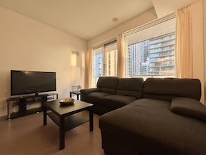 Sala de estar com televisão de tela plana, sofá escuro, mesa de centro e janela com cortinas.