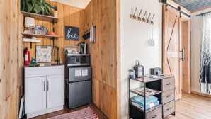 Cozinha compacta com paredes de madeira, bancada branca e refrigerador preto. Há prateleiras com plantas e utensílios de cozinha, uma luminária pendente e uma porta de celeiro deslizante.