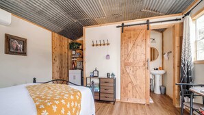 Quarto com cama, guarda-roupa de madeira, escrivaninha com cadeira, mesa pequena e banheiro com pia e espelho.