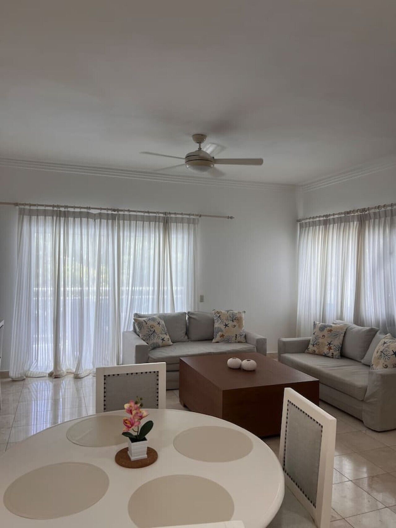 Uma sala de estar com um ventilador de teto, dois sofás com almofadas estampadas, uma mesa de centro redonda e uma mesinha com um vaso de flores.