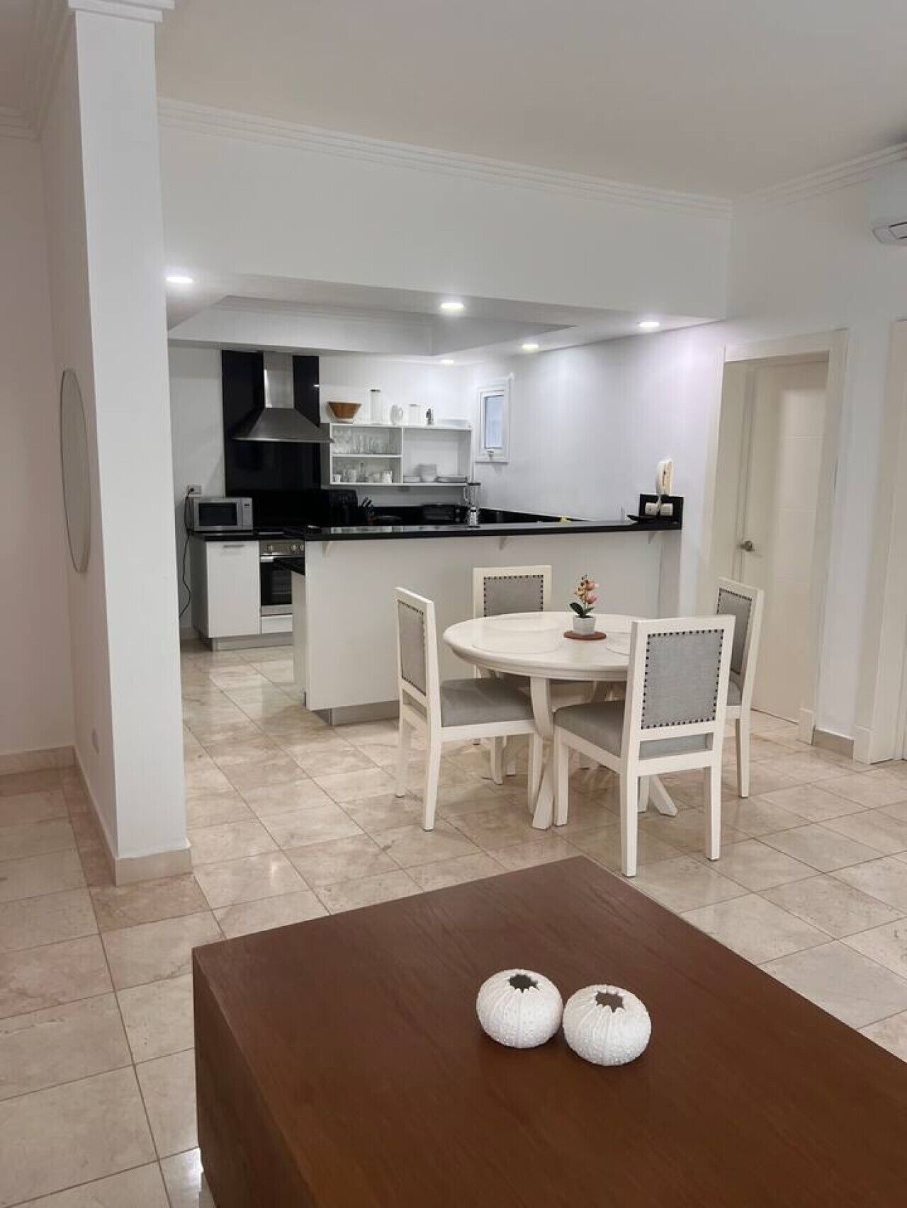 Cozinha moderna com armários brancos, sala de jantar com mesa redonda e cadeiras, e uma mesa de centro marrom com objetos decorativos.