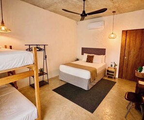 Quarto de hotel compacto com beliche, uma cama de solteiro, ventoinha no teto, mesa de cabeceira pequena com abajur e porta de madeira.