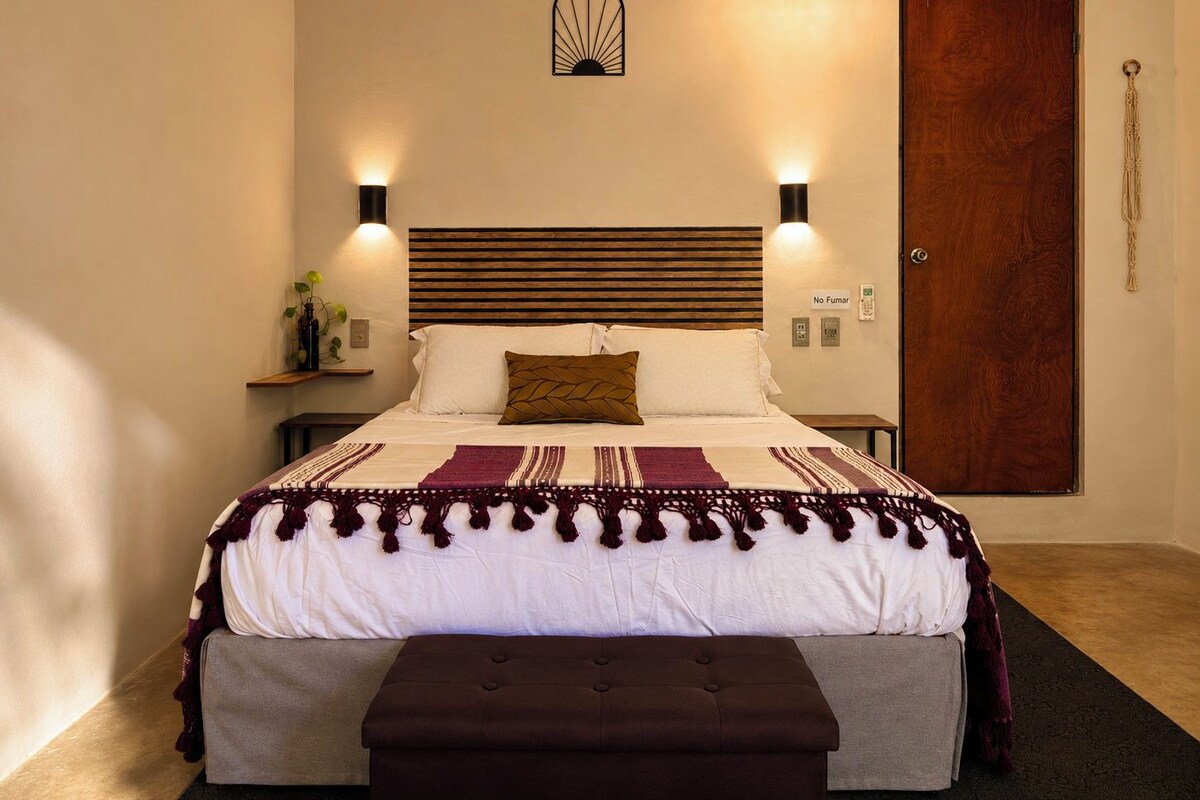 Quarto de hotel com cama grande, luminárias de cabeceira, porta de madeira e um objeto decorativo fixado na parede.