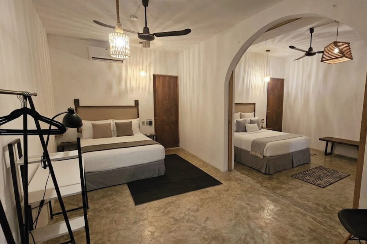 Quarto de hotel com duas camas, ventoinha no teto, luminária pendente e um banco.
