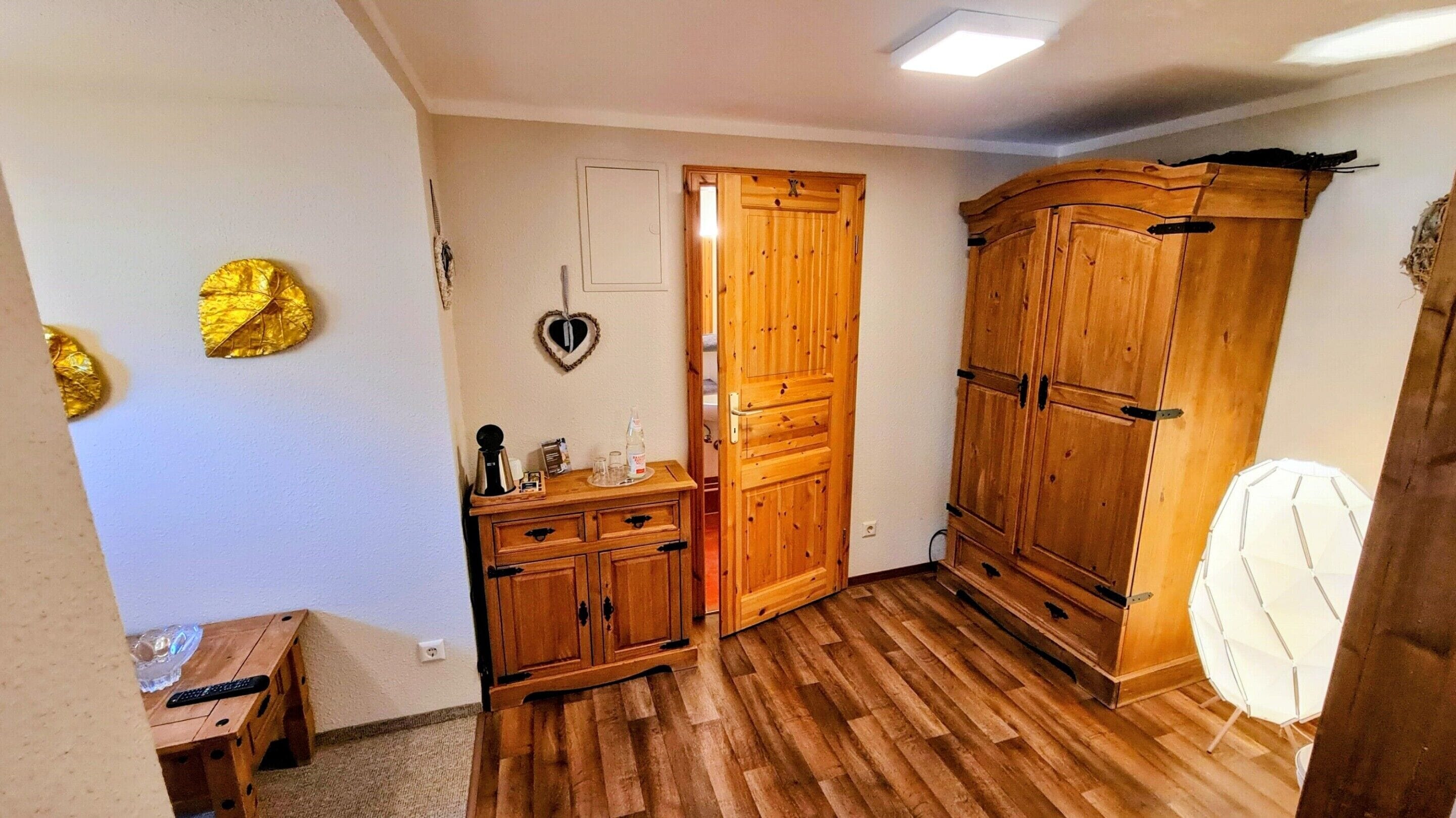 Quarto com piso de madeira, guarda-roupa de madeira, uma pequena escrivaninha de madeira e um ventilador branco.